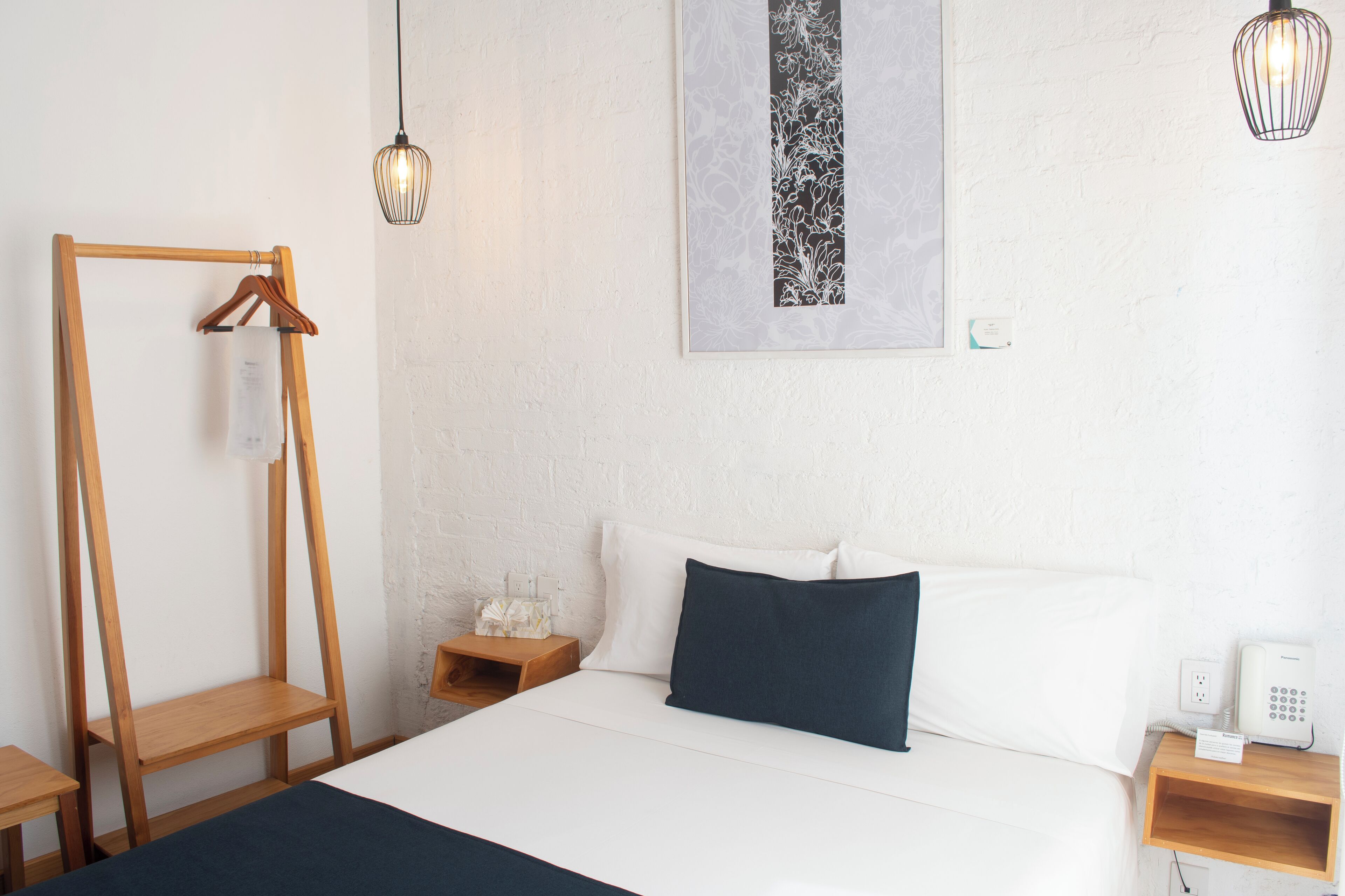 Quarto de hotel bem arrumado com cama, um cabide de madeira, mesa de cabeceira com telefone e luminária fixada na parede.