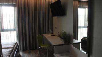 Quarto de hotel moderno com cama, escrivaninha, cadeira, TV fixada na parede e janela com cortinas.