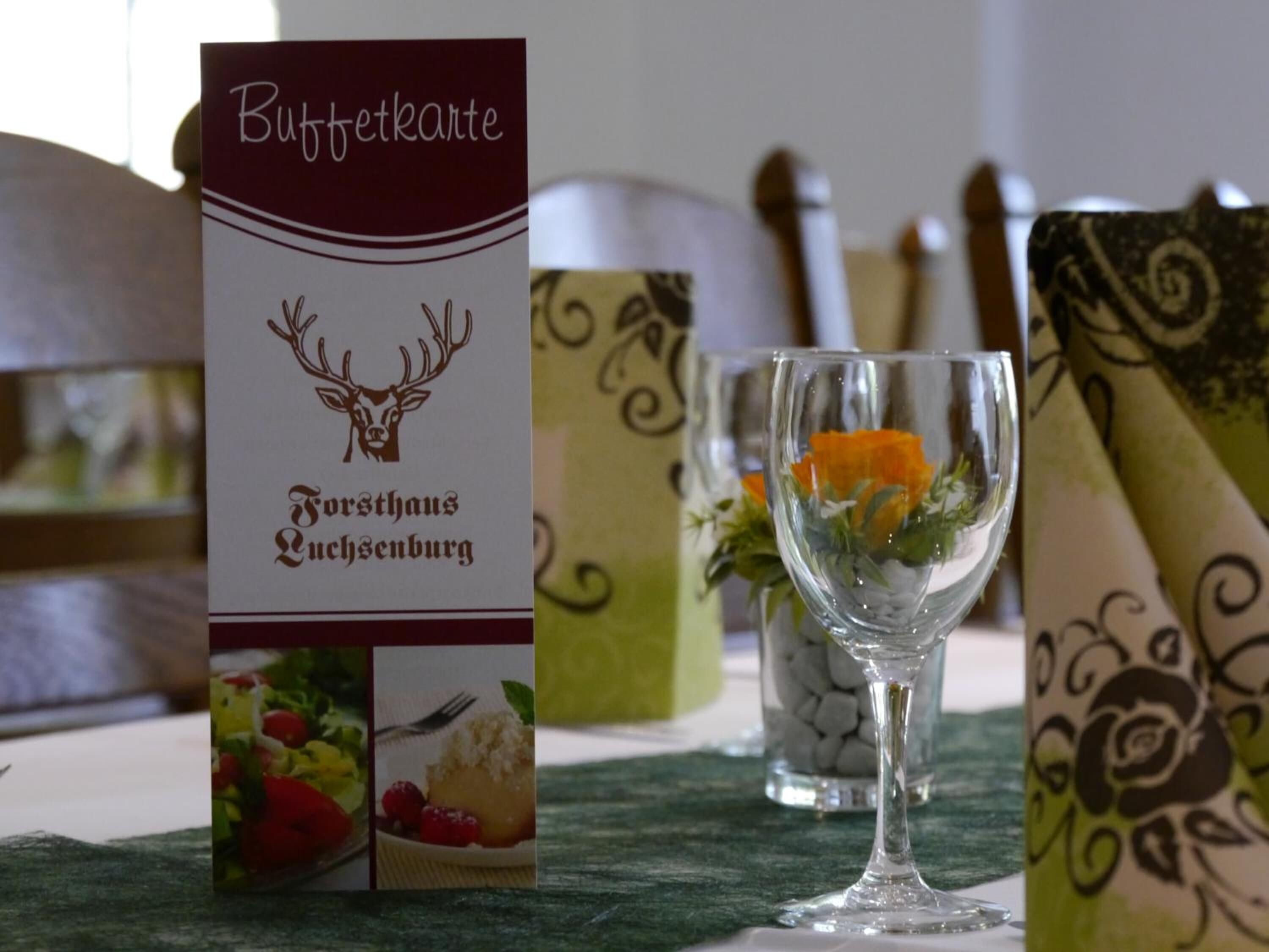 Um menu intitulado "Buffetkarte" com um logotipo de veado e o texto "Forsthaus Luchsenburg", um copo com uma fatia de laranja e folhagens, e toalhas de mesa estampadas.