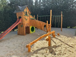 Estrutura de madeira para brincar com escorrega e elementos de escalada, sobre o chão de areia, com árvores ao fundo.