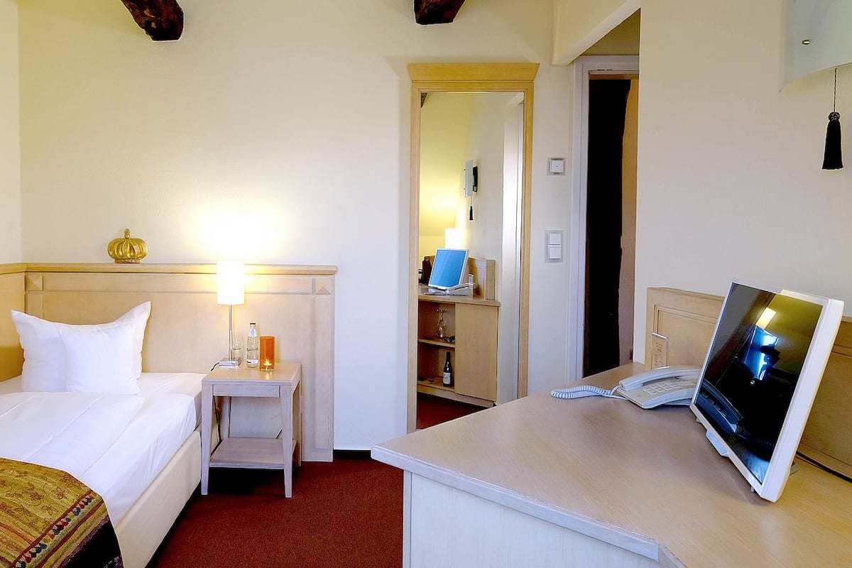 Quarto de hotel com cama, mesa de cabeceira, luminária e escrivaninha com monitor de computador.