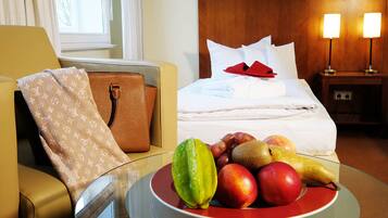 Quarto de hotel com cama, mesa de cabeceira com abajur, cadeira e mesa de vidro com frutas.