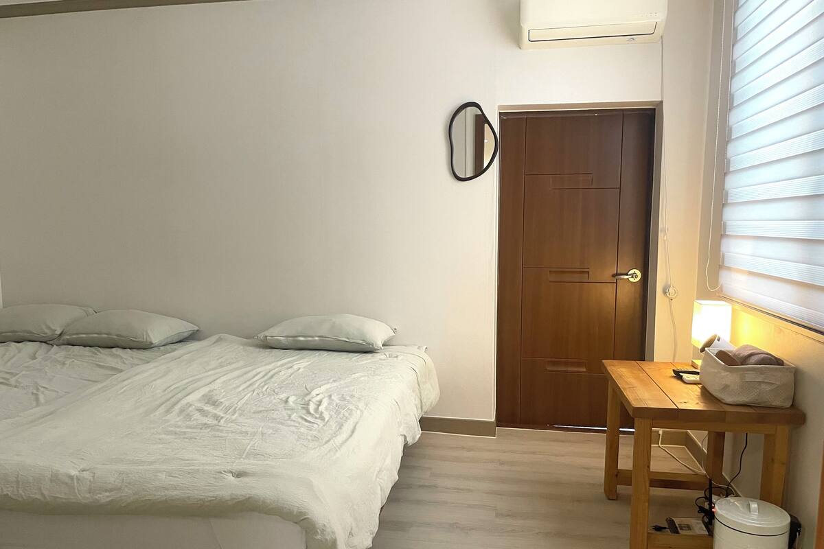 Quarto com cama, mesa de cabeceira com abajur, criado-mudo de madeira e lixeira branca.