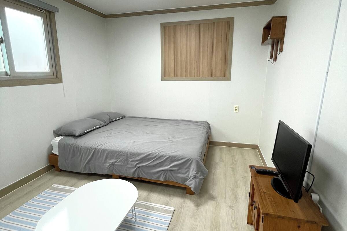 Quarto com cama, televisão sobre um suporte de madeira, janela e luminária no teto.