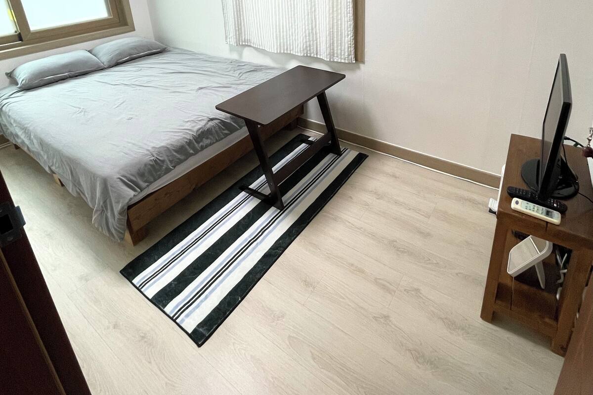 Quarto com cama, mesinha, aparador para TV, janela com cortinas e luminária no teto.
