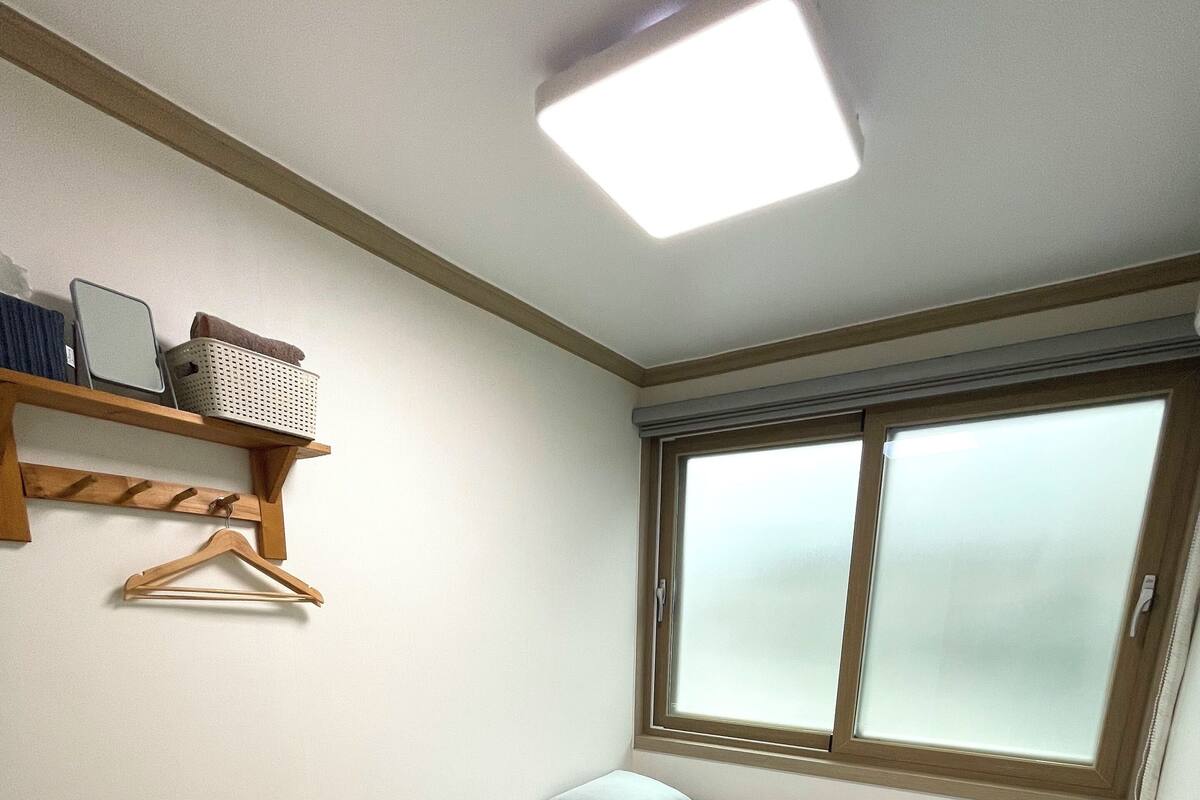 Um quarto pequeno e bem organizado, com uma cama, uma escrivaninha, uma cadeira, uma janela e uma prateleira com um cesto.