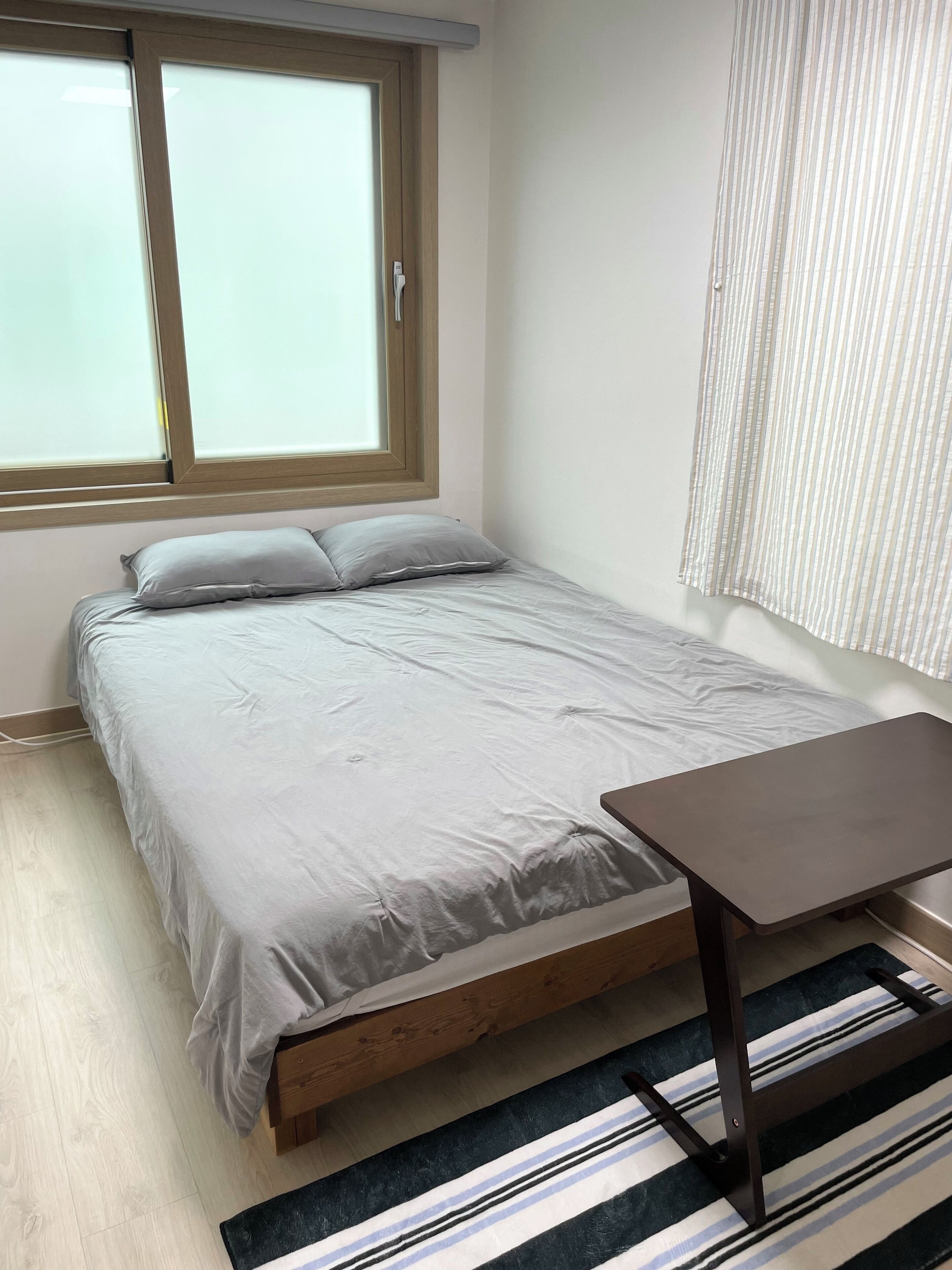 Quarto com cama, mesa de cabeceira de madeira, janela com persianas e tapete listrado.
