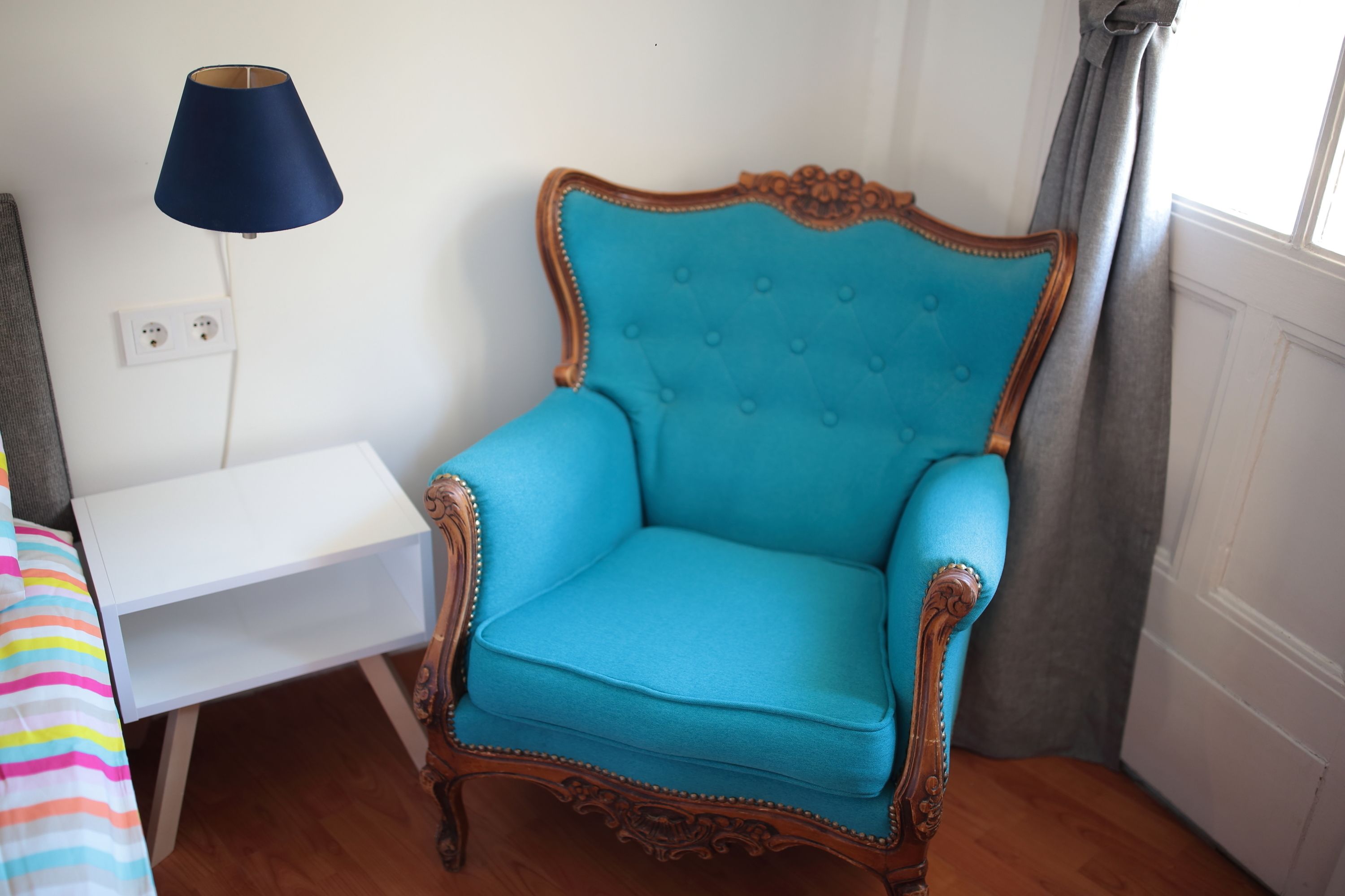 Quarto com poltrona estofada azul, mesa de apoio branca, luminária e janela com cortinas.