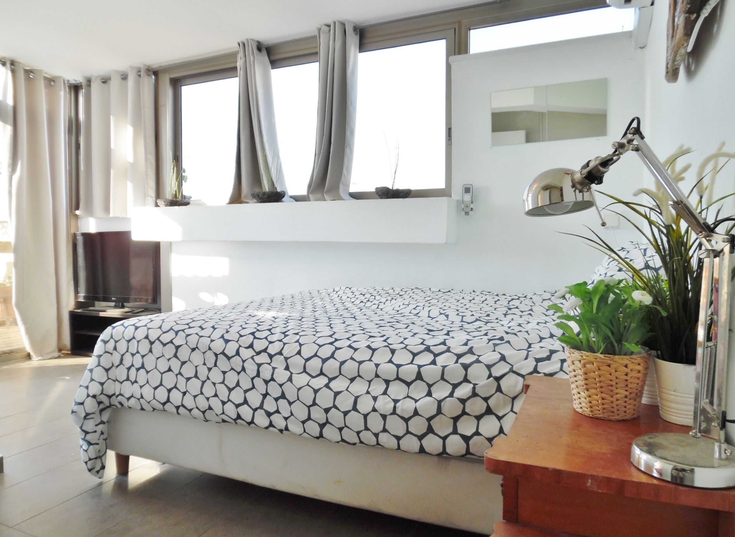 Quarto com cama, escrivaninha com luminária, televisão e janela com cortinas.