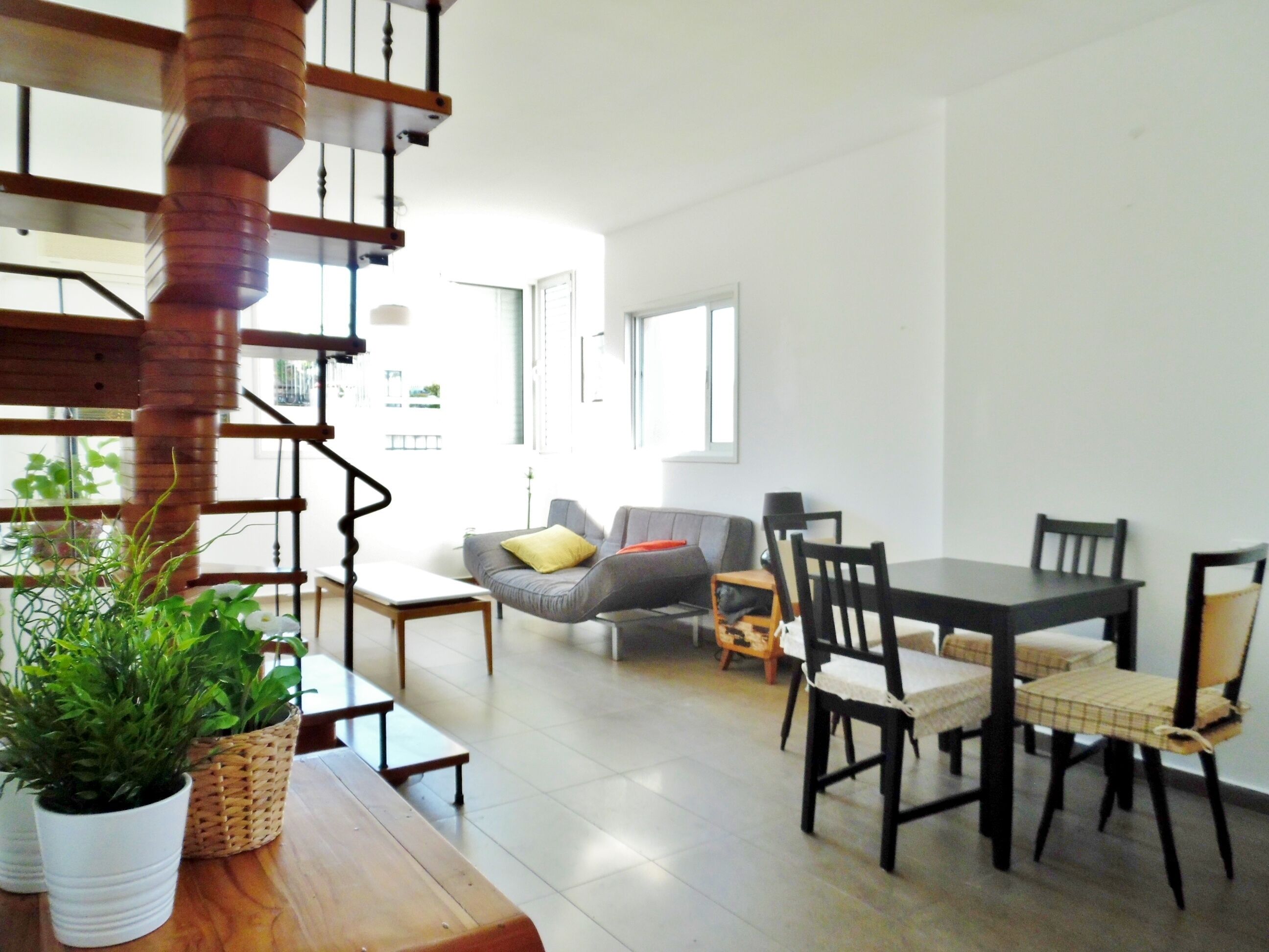 Um espaço de estar moderno com escada, área de jantar com mesa e cadeiras, sofá com almofadas e um vaso de planta.