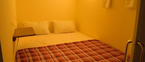 Quarto pequeno com cama de solteiro, colcha xadrez, mesa de cabeceira e um porta-toalhas.