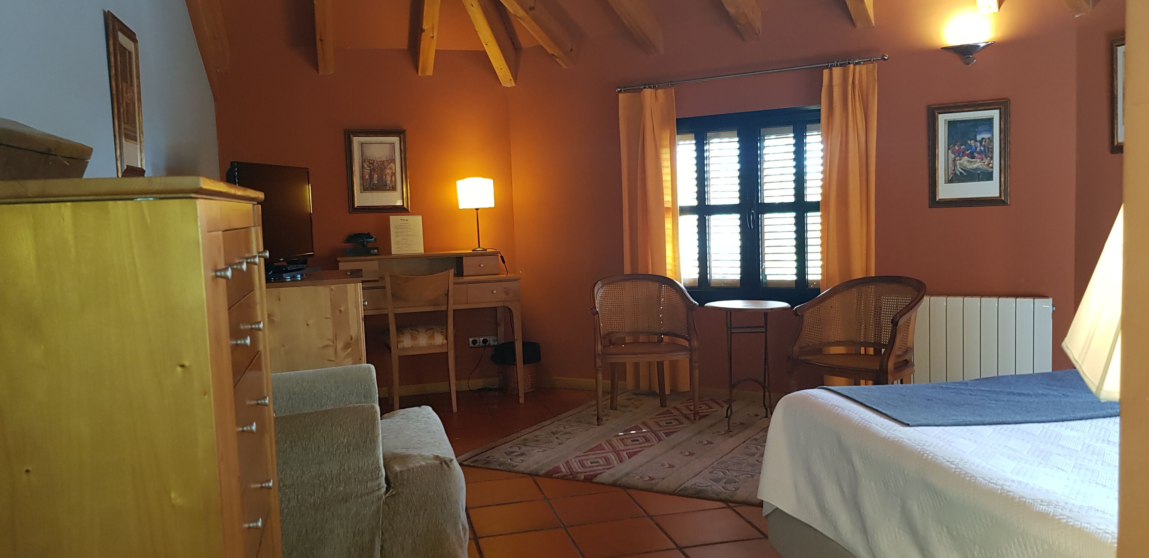 Um quarto aconchegante com uma cama, uma escrivaninha, duas cadeiras, um abajur e uma janela com cortinas.