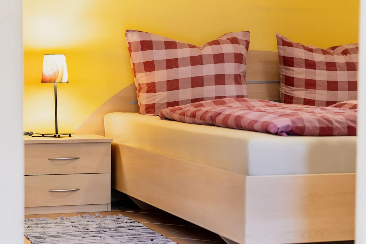 Cama bem arrumada com colcha xadrez, abajur e mesa de cabeceira de madeira com gavetas.