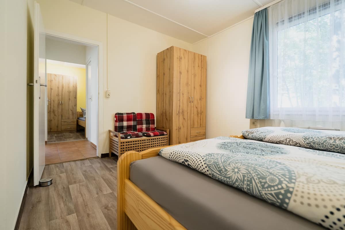 Quarto com cama, guarda-roupa de madeira, um cantinho de estar com manta xadrez e vista para o exterior pela janela.