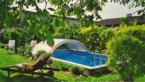 Uma piscina azul com toldo, rodeada por vegetação exuberante e um prédio branco ao fundo.