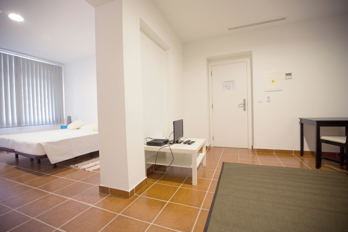 Quarto de hotel com uma cama, uma mesa com um computador, uma cadeira e uma porta.