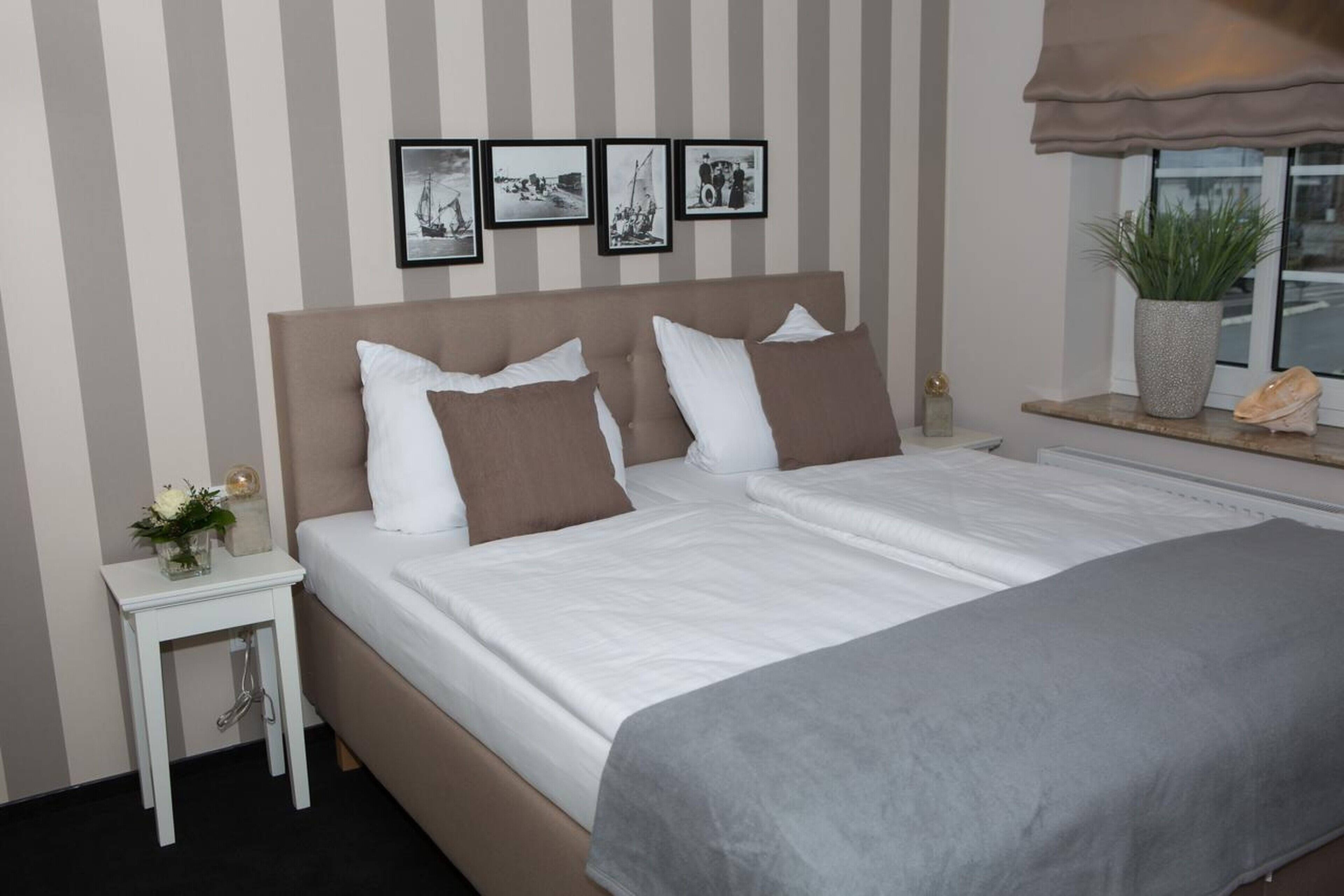 Cama bem arrumada com roupa de cama branca e castanha, uma mesinha de cabeceira com um vaso de flores e uma janela com vista para a rua.