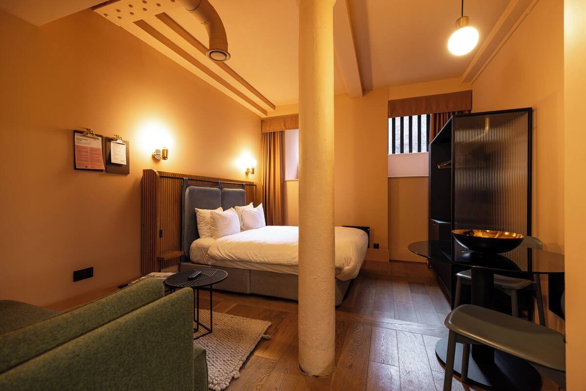 Quarto de hotel com cama, escrivaninha, cadeira, mesa pequena e um grande pilar.