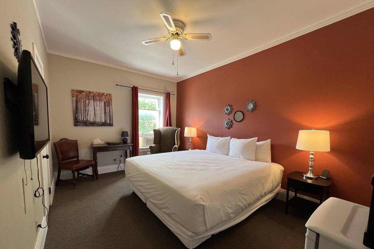 Quarto de hotel com uma cama grande, duas mesas de cabeceira com abajures, uma escrivaninha com cadeira, um ventilador de teto e uma janela com cortinas.