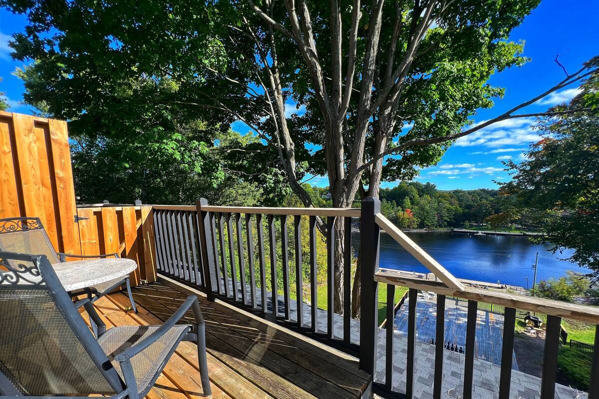 Um deck de madeira com guarda-corpo de metal, vista para um lago, um banco e uma mesa com cadeiras.