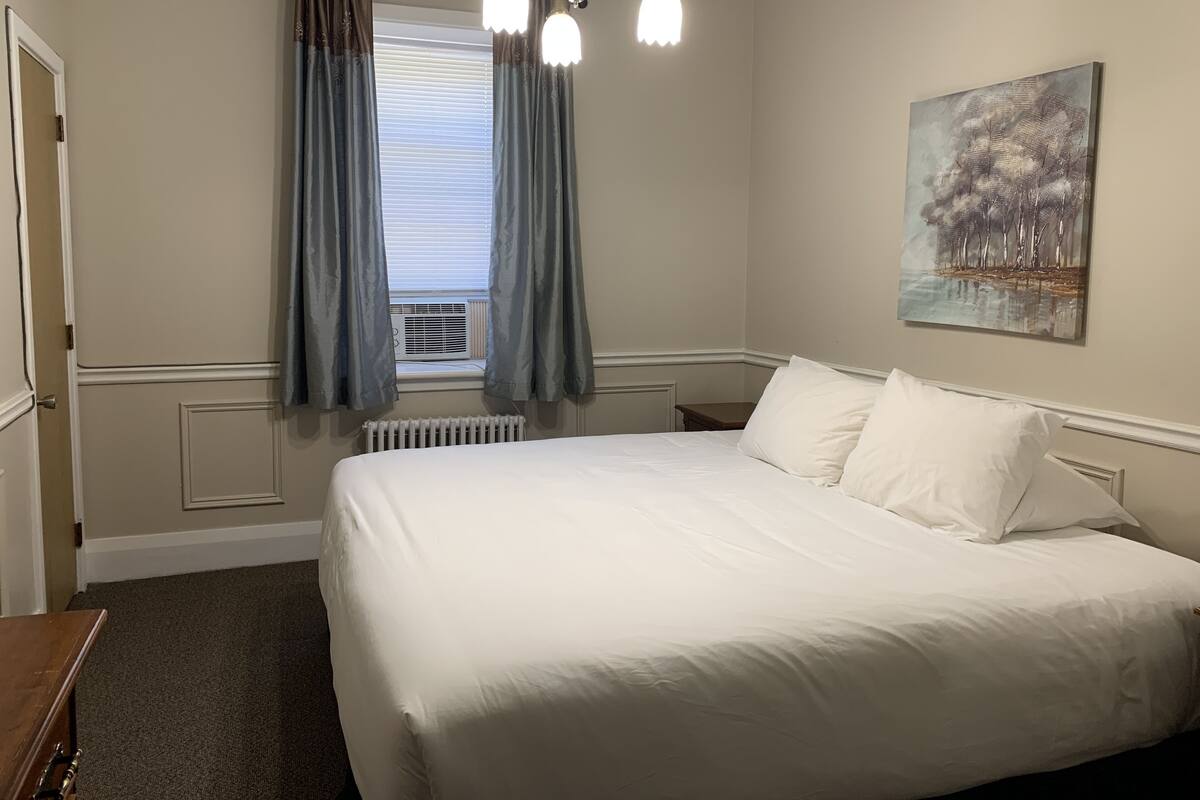 Quarto com cama grande, radiador, janela com cortinas e um quadro na parede.