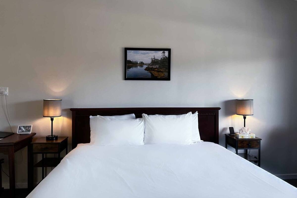 Uma cama bem arrumada com roupa de cama branca, duas mesas de cabeceira com abajures e um quadro com paisagem na parede.