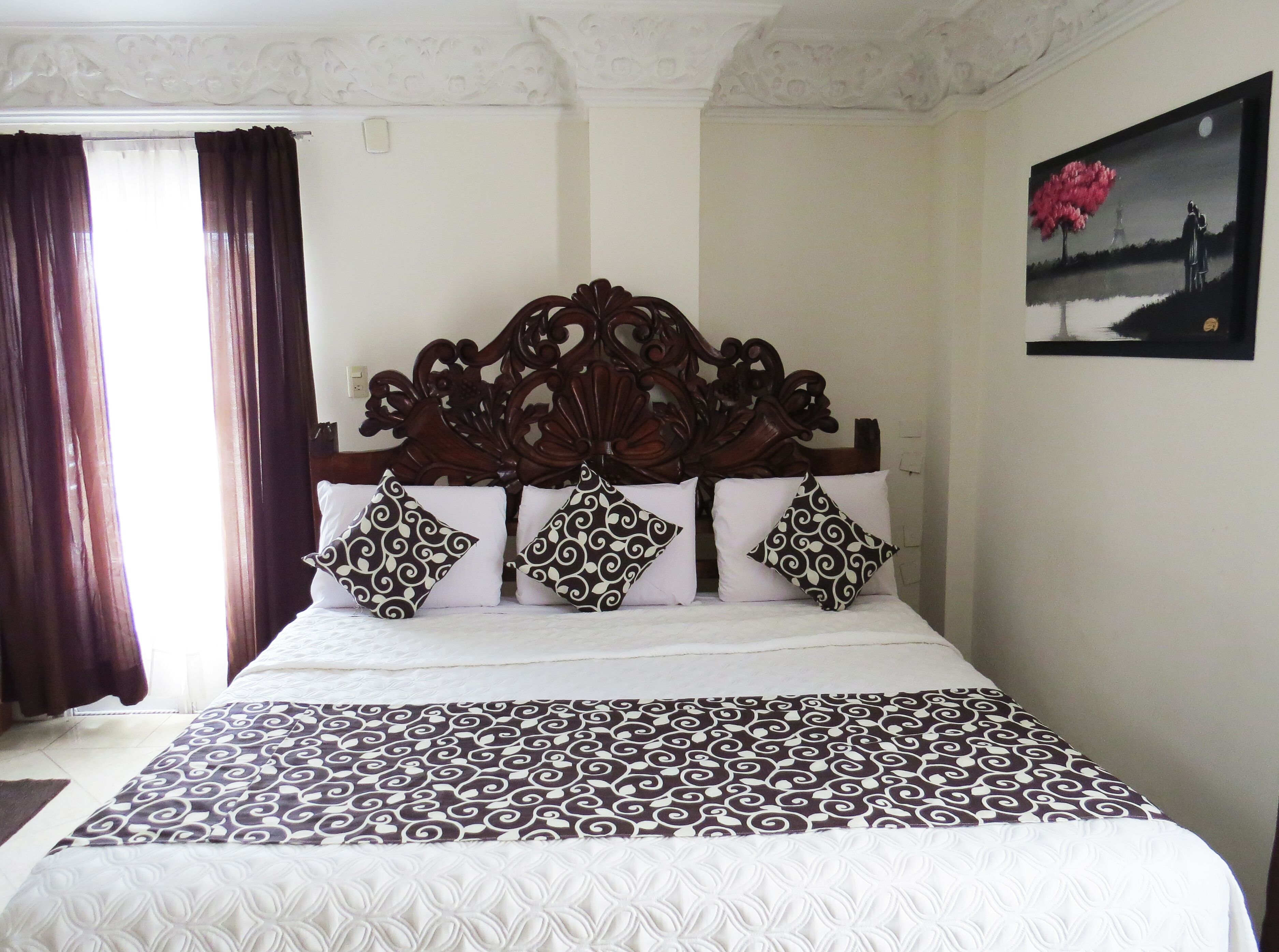 Quarto de hotel com uma cama grande, cabeceira de madeira ornamentada, roupa de cama estampada e um quadro na parede.