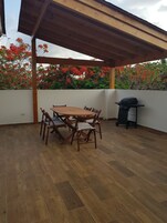 Alpendre coberto com mesa e cadeiras de madeira, churrasqueira e vista para flores vermelhas.