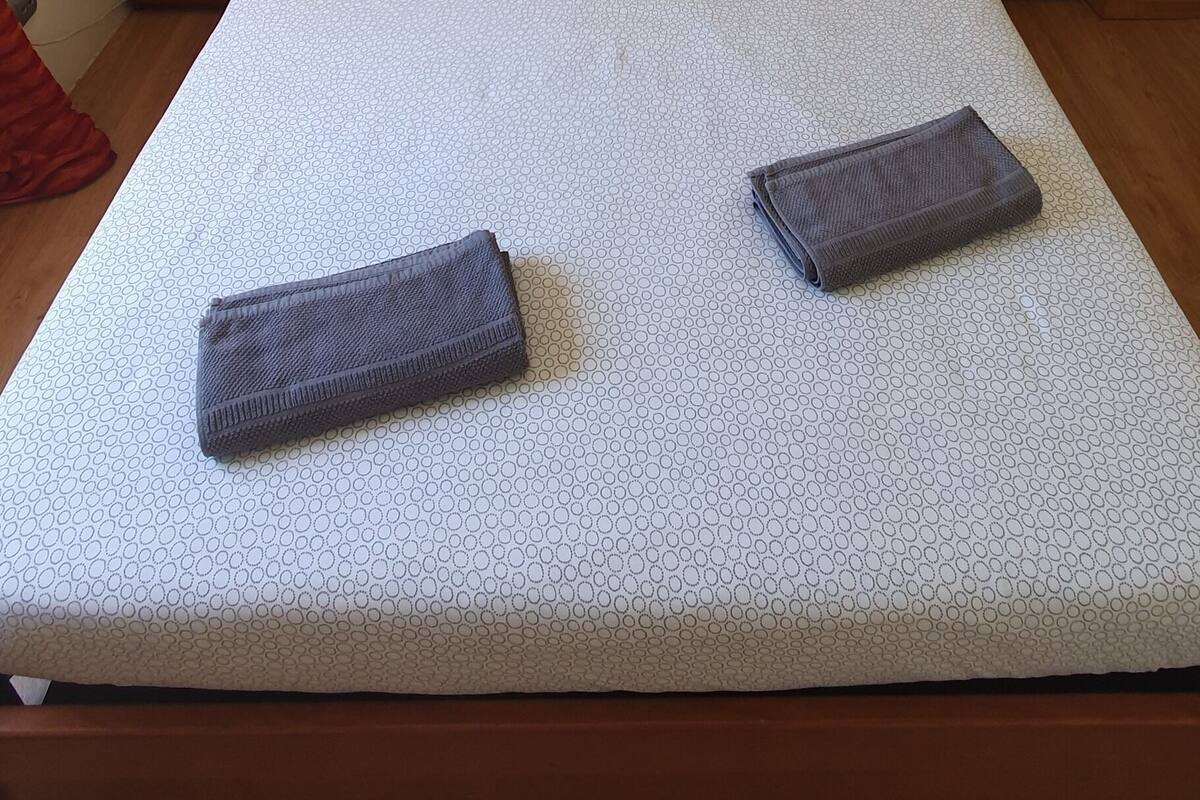 Cama bem arrumada com um edredom estampado, dois travesseiros e duas toalhas dobradas. Duas mesas de cabeceira, com luminárias e um controle remoto em uma delas.