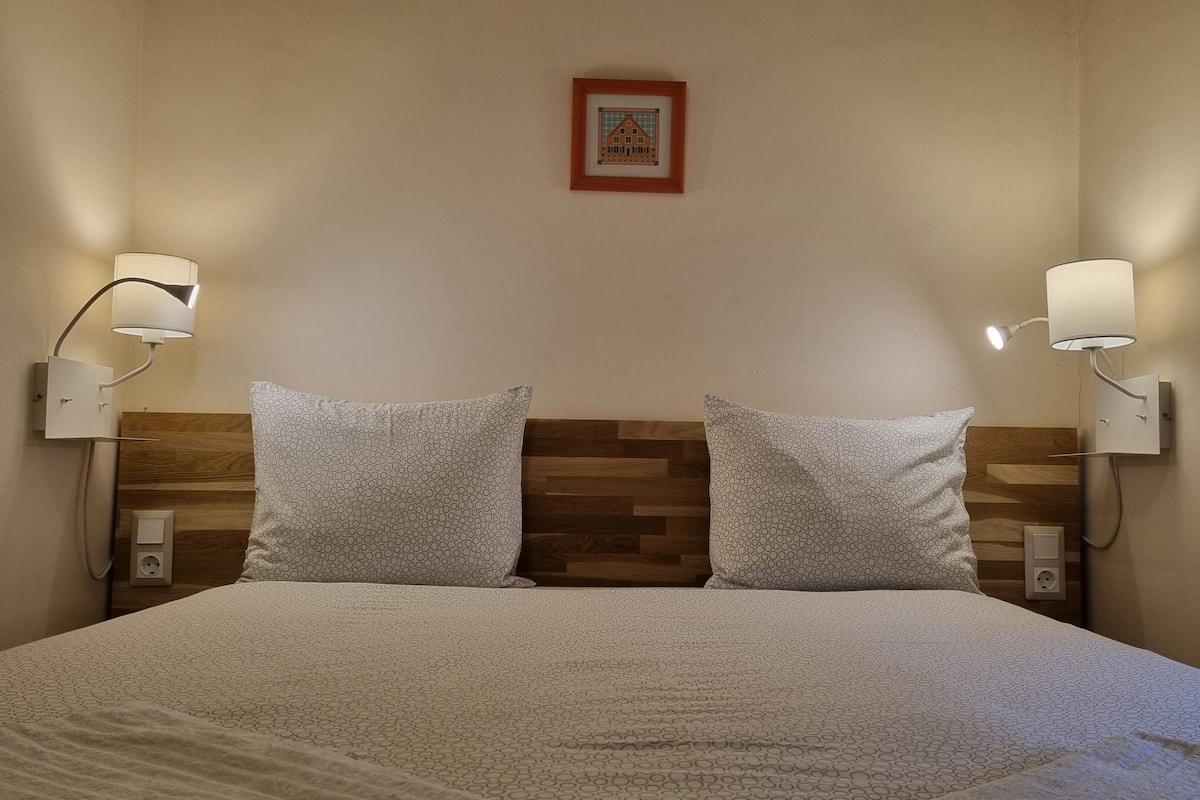 Cama bem arrumada com colcha estampada e duas almofadas, com luminárias fixadas na parede e um quadro emoldurado acima.