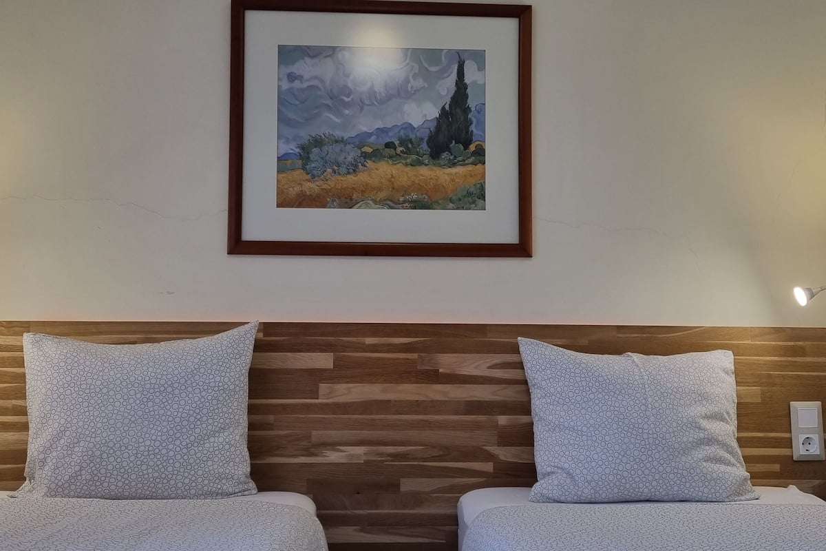 Quarto de hotel com duas camas, uma cabeceira de madeira, um quadro emoldurado acima e luminárias fixadas na parede.