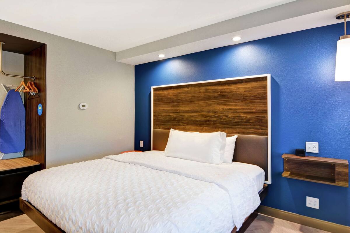 Quarto de hotel moderno com uma cama grande, cabeceira de madeira, parede azul e uma pequena prateleira de madeira.