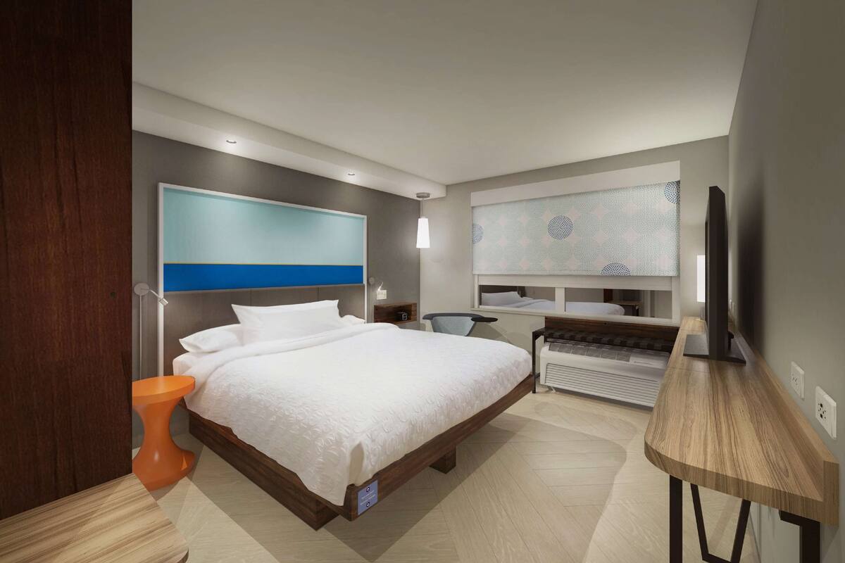 Quarto de hotel moderno com uma cama grande, uma escrivaninha, uma cadeira, televisão e uma obra de arte na parede.