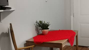 Quarto com cadeira de madeira, pequena mesa com um tecido vermelho, planta em vaso, televisão em uma estante e uma porta branca.