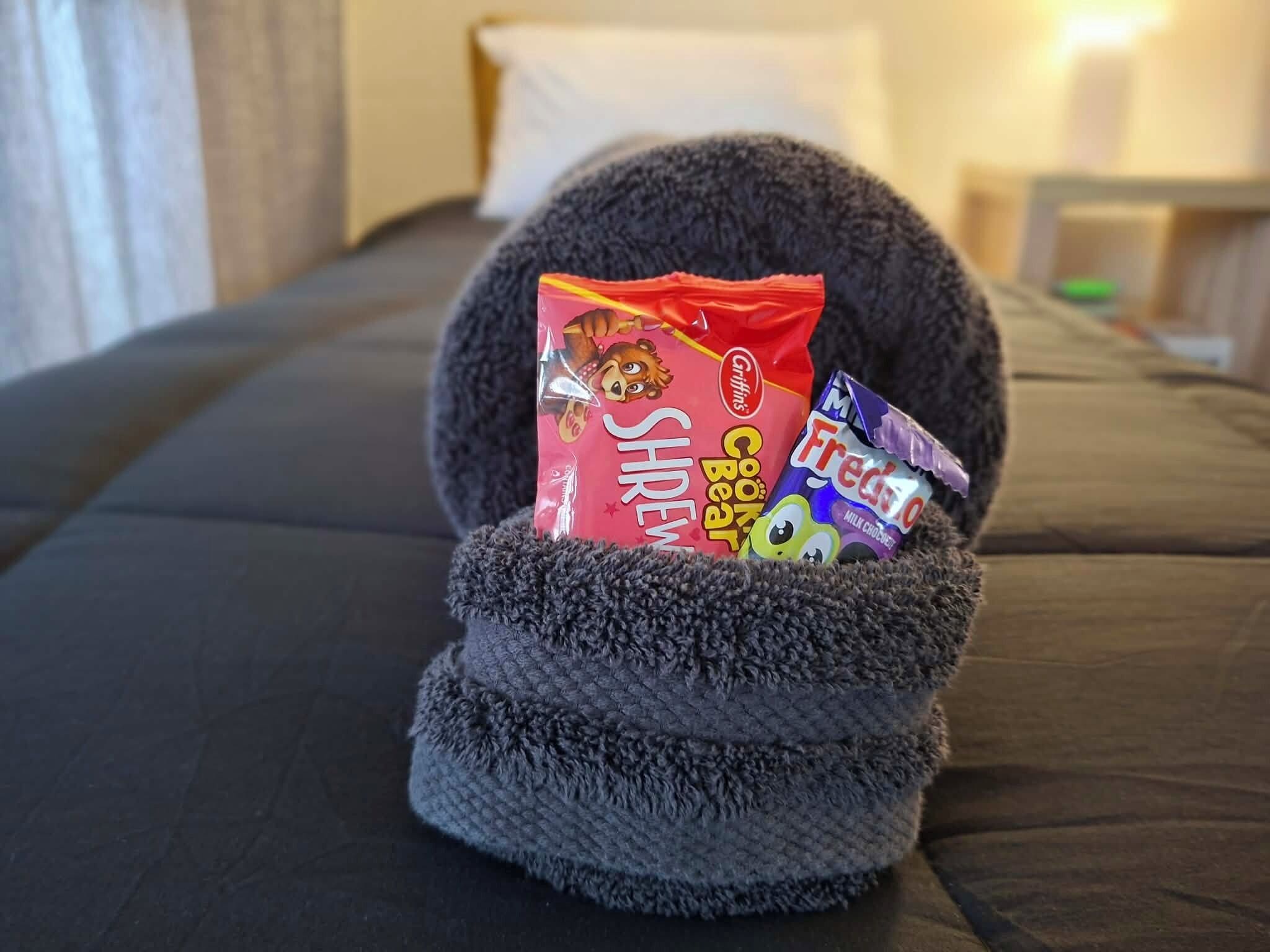 Um objeto macio e redondo cinzento sobre uma cama, com um travesseiro e uma mesinha de cabeceira ao fundo.