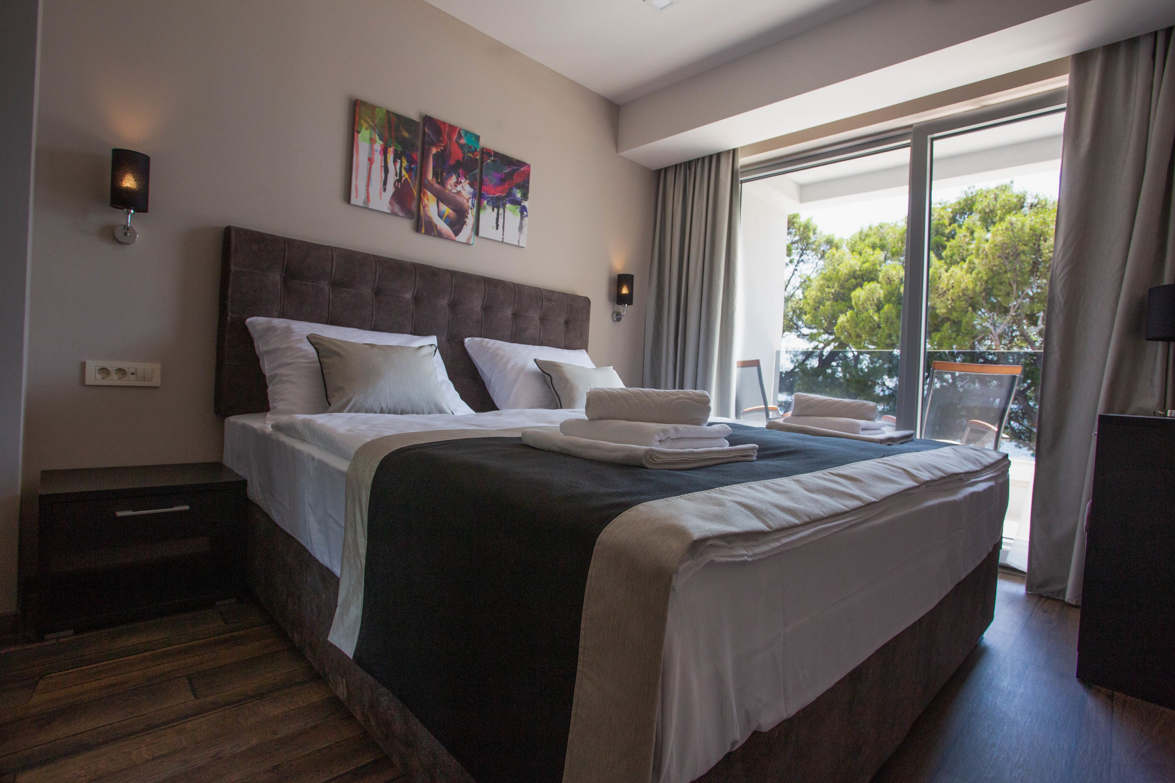 Quarto de hotel moderno com uma cama grande, mesa de cabeceira, janela com cortinas e uma obra de arte na parede.