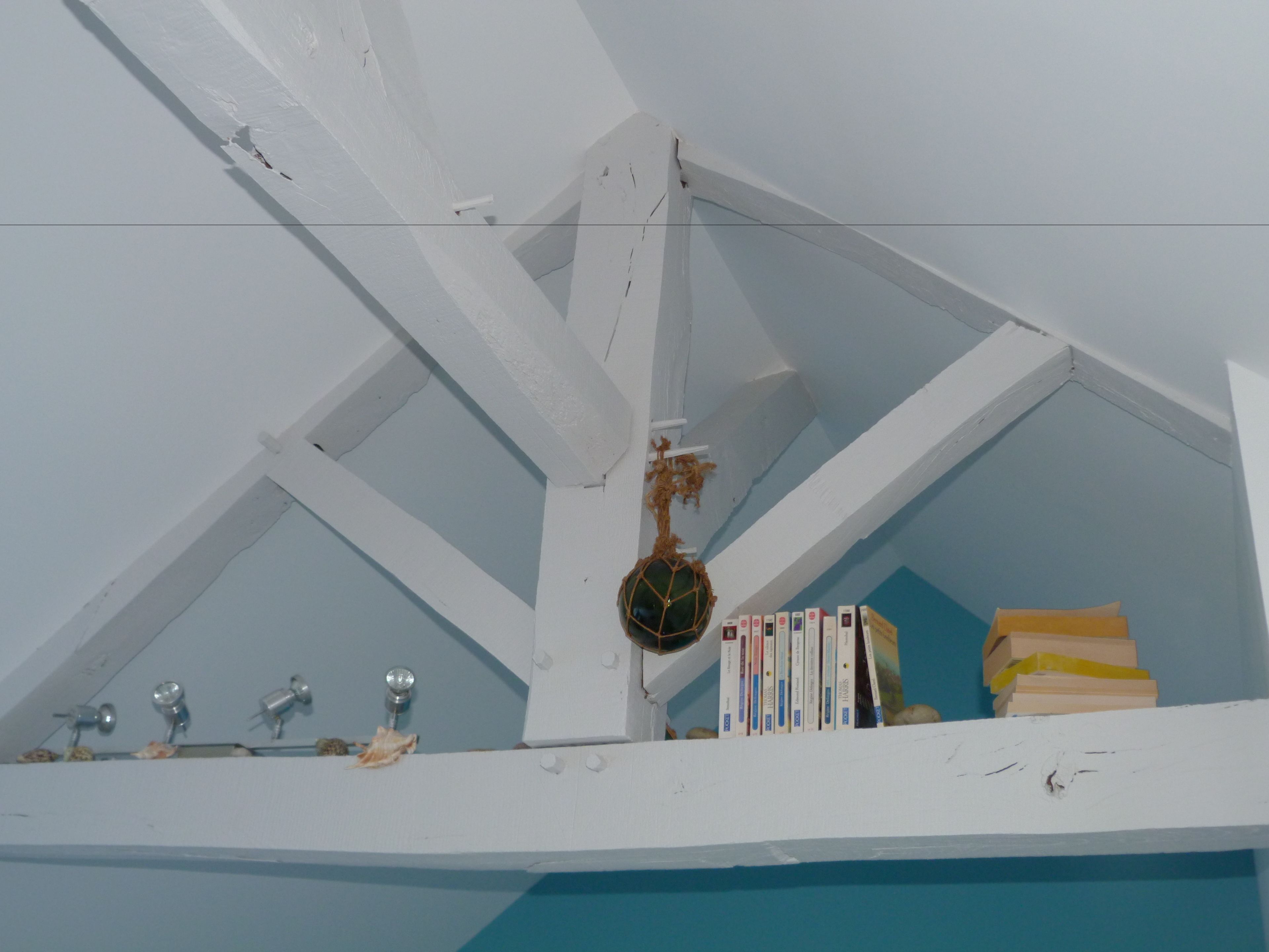 Teto com vigas brancas, um adorno pendurado e uma estante com livros e objetos decorativos.