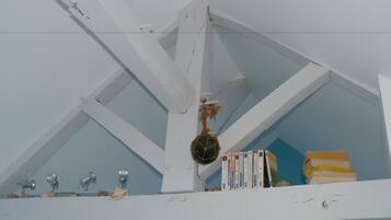 Teto com vigas brancas, um adorno pendurado e uma estante com livros e objetos decorativos.