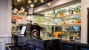 Uma área de bar com balcão de vidro, prateleiras com garrafas de bebidas e uma placa indicando a idade mínima exigida.