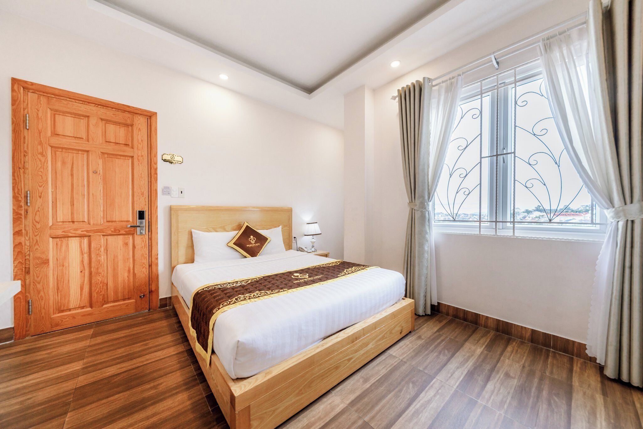 Quarto de hotel com cama de madeira, uma janela grande com cortinas e porta de madeira.