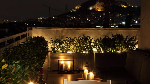 Terraço no telhado com vista para a cidade à noite, com área de estar com almofadas e uma mesa pequena, rodeada por vasos de plantas e velas.