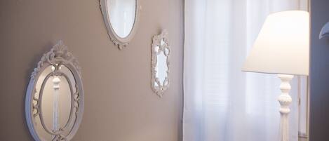 Quarto com três espelhos ornamentados na parede, uma poltrona estofada, um abajur branco e uma janela com cortina translúcida.