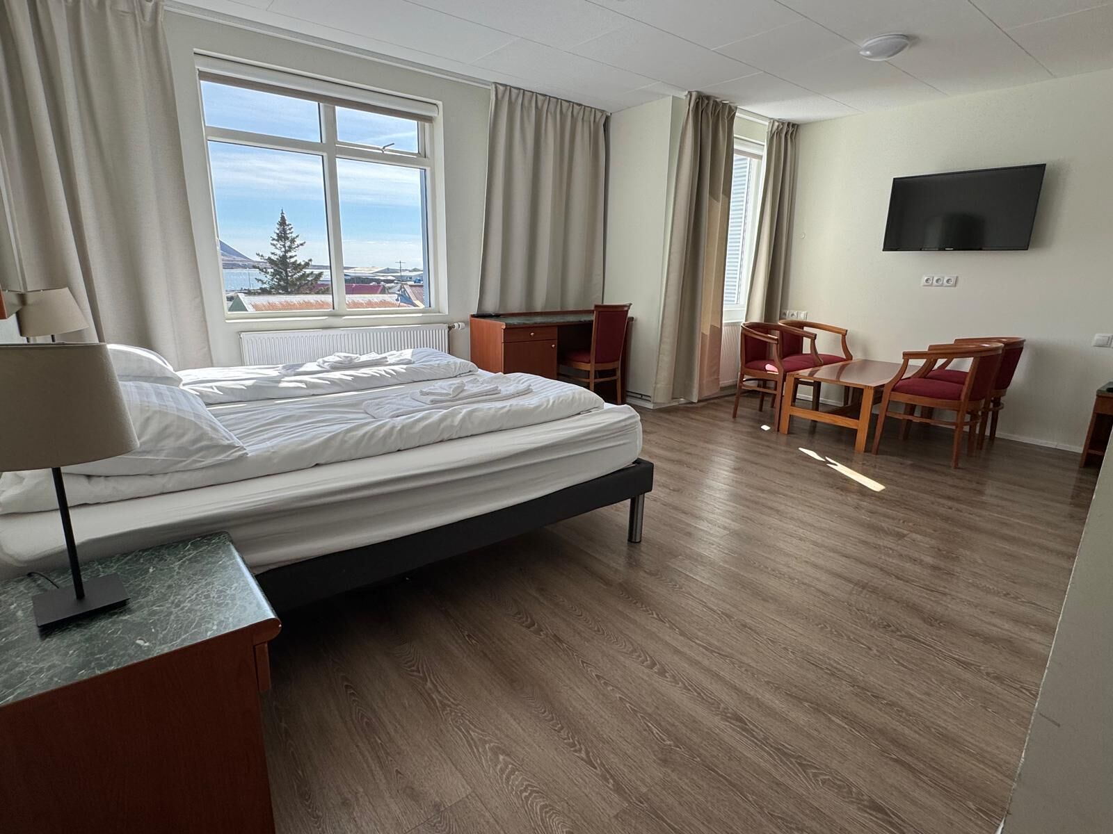 Quarto de hotel com uma cama, uma escrivaninha, uma cadeira, uma TV e uma janela com cortinas.