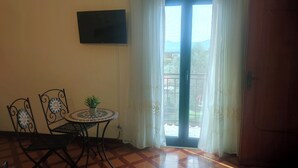 Quarto com uma pequena mesa, duas cadeiras, um vaso de planta, uma TV na parede e vista para uma varanda com porta.