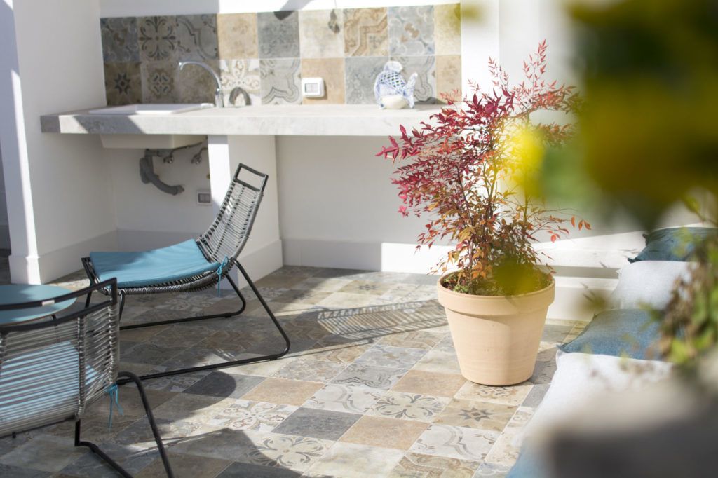 Um pátio banhado pelo sol, com piso de azulejos, pia branca, cadeira azul e um vaso com planta.
