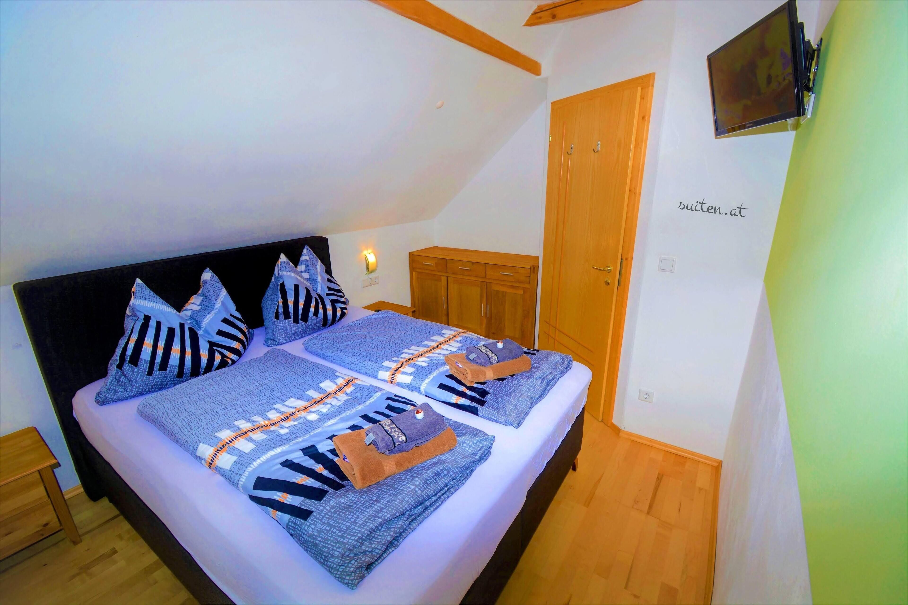 Cama de casal com cabeceira, duas almofadas, um cobertor e uma toalha dobrada. Dispõe de um guarda-roupa de madeira e uma porta com um aviso em cima.