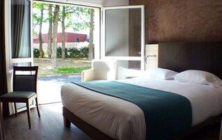 Quarto de hotel com cama, uma cadeira e uma janela grande com vista para árvores e um prédio lá fora.