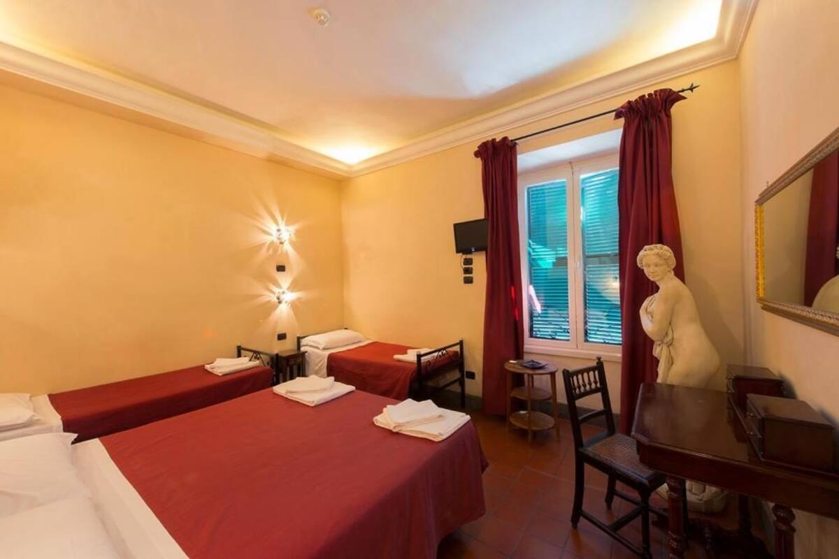 Quarto de hotel com duas camas de solteiro, uma escrivaninha, uma cadeira, uma estátua e uma janela com cortinas.