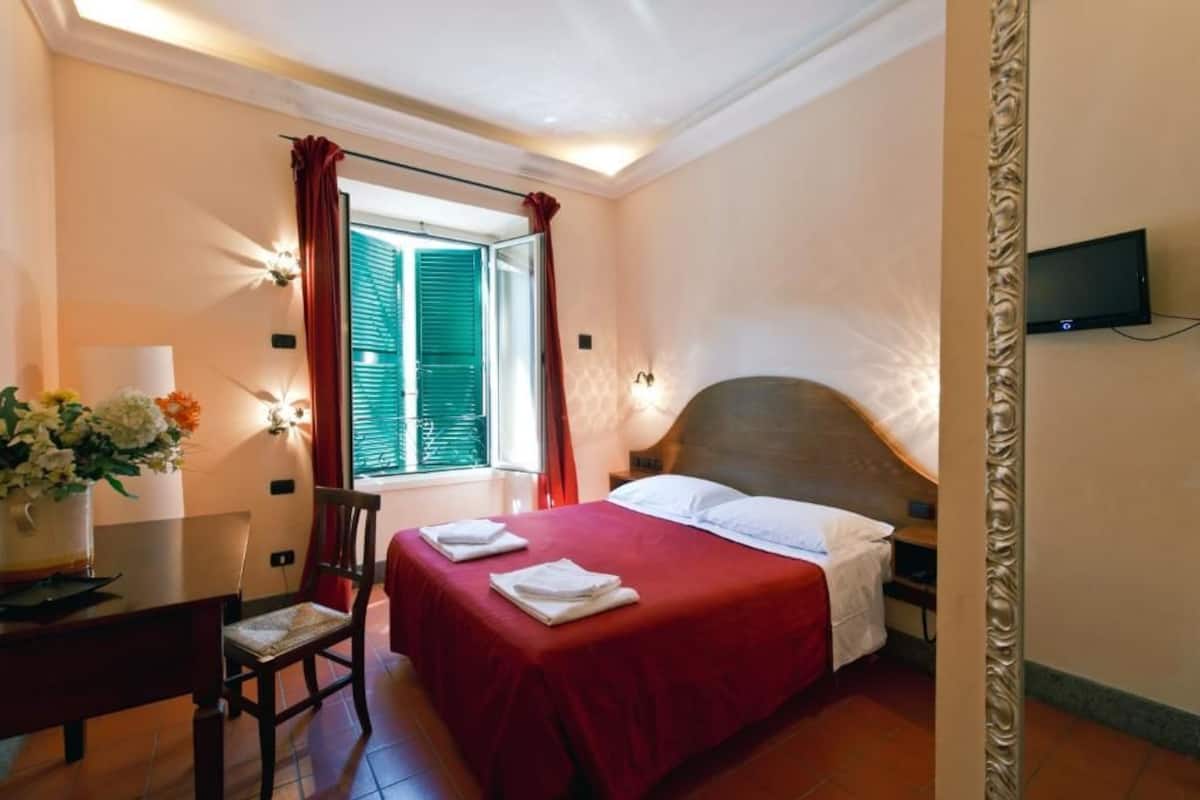 Quarto de hotel com cama, escrivaninha com cadeira, janela com cortinas e televisão de tela plana na parede.