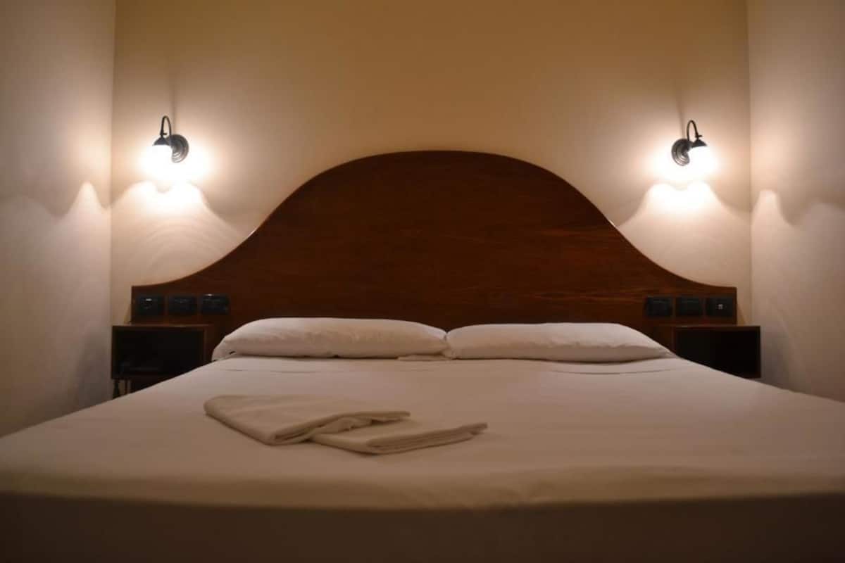 Cama bem arrumada com lençóis brancos e toalha dobrada, com duas mesas de cabeceira com luminárias, em quarto com cabeceira de madeira e paredes em tons claros.